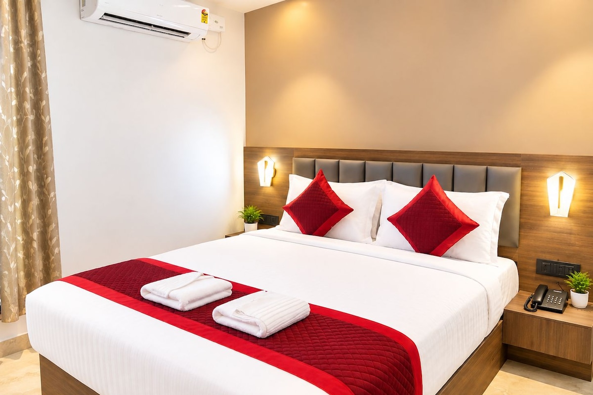 Quarto de hotel com uma cama, mesa de cabeceira com telefone e uma planta, e um aparelho de ar condicionado na parede.