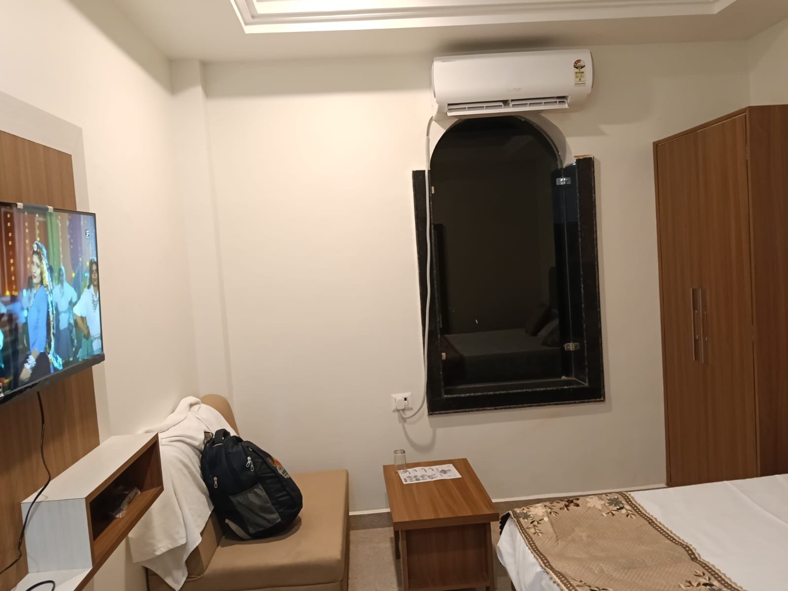 Quarto de hotel com cama, televisão fixada na parede, guarda-roupa de madeira, mesa pequena e aparelho de ar condicionado.
