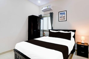 Quarto com cama, mesa de cabeceira, abajur, ar condicionado, janela com cortinas e quadro pendurado na parede.