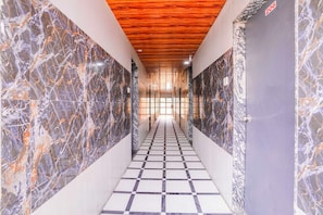Corredor com paredes de mármore e chão xadrez, que leva a um espaço interior iluminado.