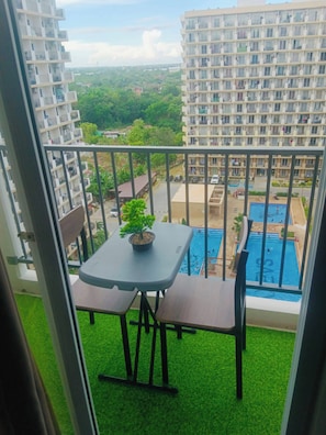 Uma varanda com mesa e cadeiras, um pequeno vaso de planta e vista para uma piscina e edifícios altos.