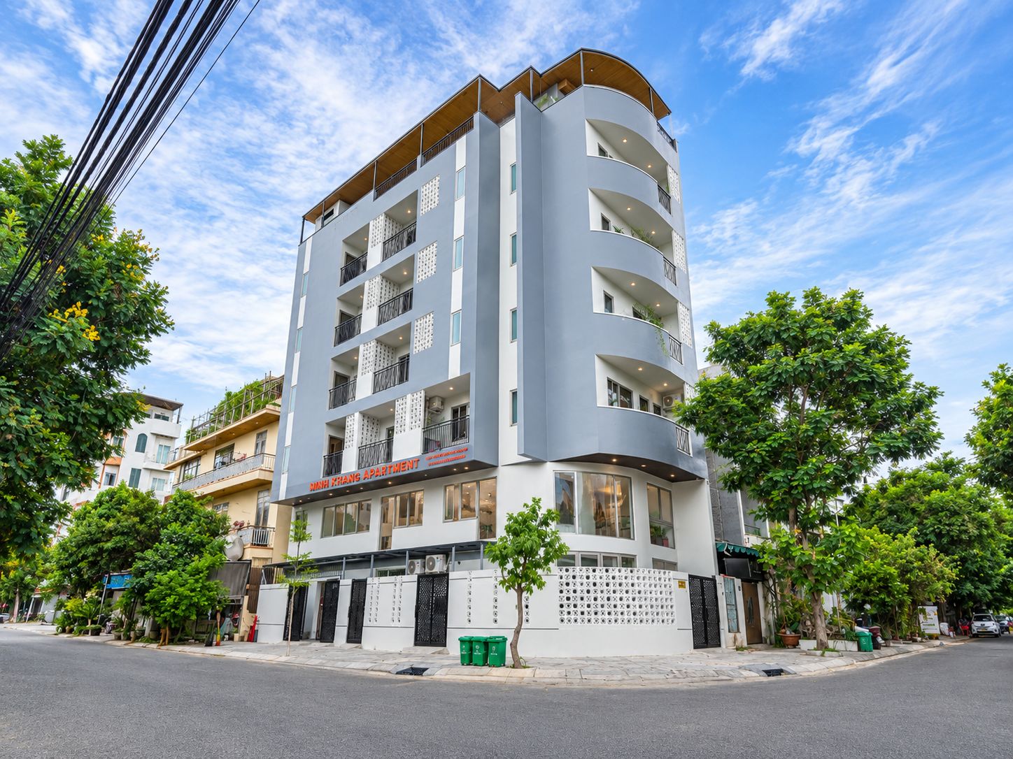 Um edifício de vários andares com varandas, um letreiro indicando 'SING KHANG APARTMENT' e uma vista da rua com árvores e outros edifícios.