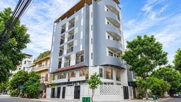 Um edifício de vários andares com varandas, um letreiro indicando 'SING KHANG APARTMENT' e uma vista da rua com árvores e outros edifícios.