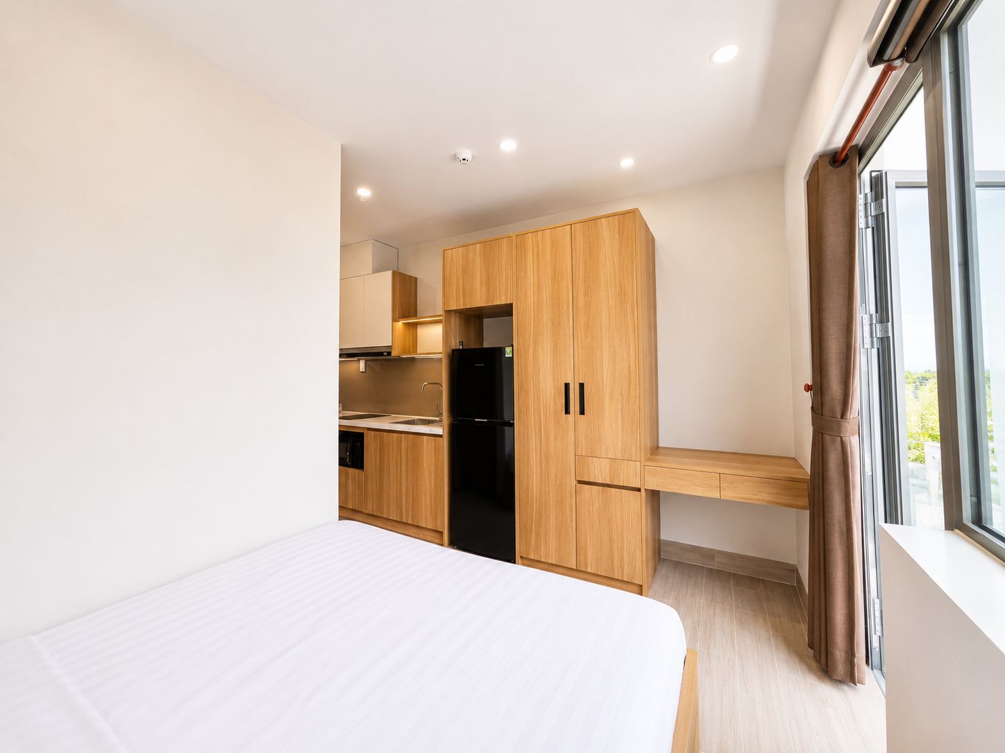 Quarto moderno com cama, guarda-roupa de madeira e área de cozinha visível por uma porta aberta.
