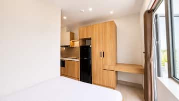 Quarto moderno com cama, guarda-roupa de madeira e área de cozinha visível por uma porta aberta.