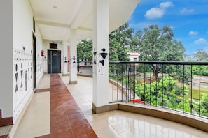 Corredor com piso ladrilhado que leva a uma varanda com grade de metal, com vista para um jardim.