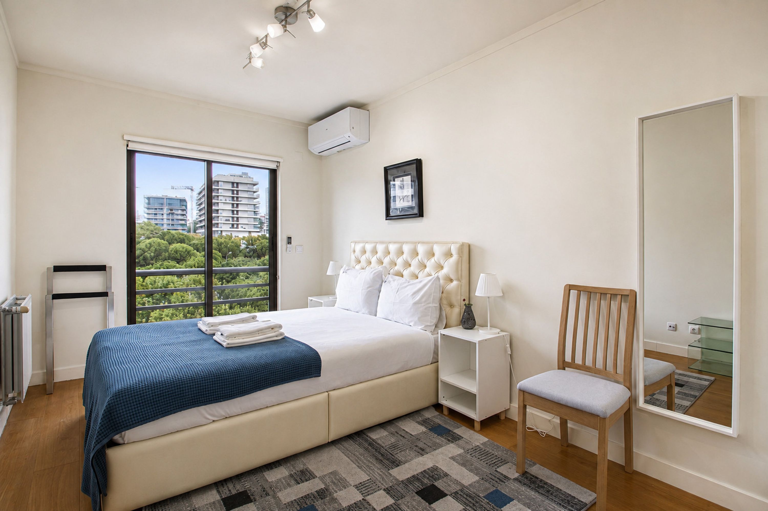 Quarto com cama grande, mesa de cabeceira, cadeira, espelho e janela com vista para prédios e árvores.
