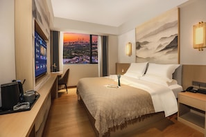 Quarto de hotel com cama, escrivaninha, cadeira, televisão e janela com vista para a cidade.