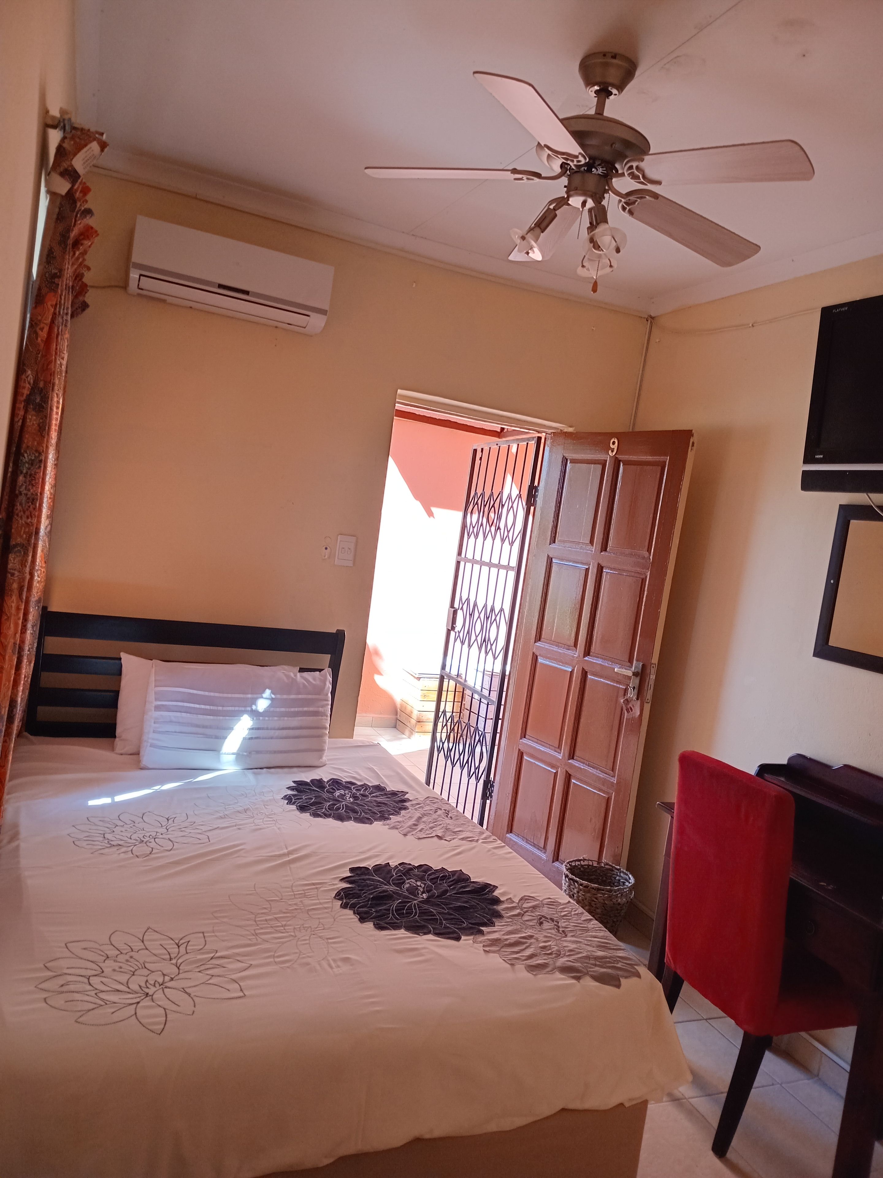 Quarto com cama, ventilador de teto, ar-condicionado, uma cadeira vermelha e televisão na parede.