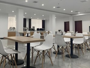 Uma zona de refeições moderna, com várias mesas e cadeiras, uma cozinha ao fundo, e um design limpo e minimalista.
