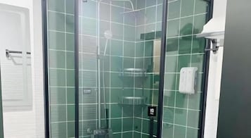 Área de chuveiro com paredes de vidro, painel de controle, dispensador de toalhas e secador de mãos.