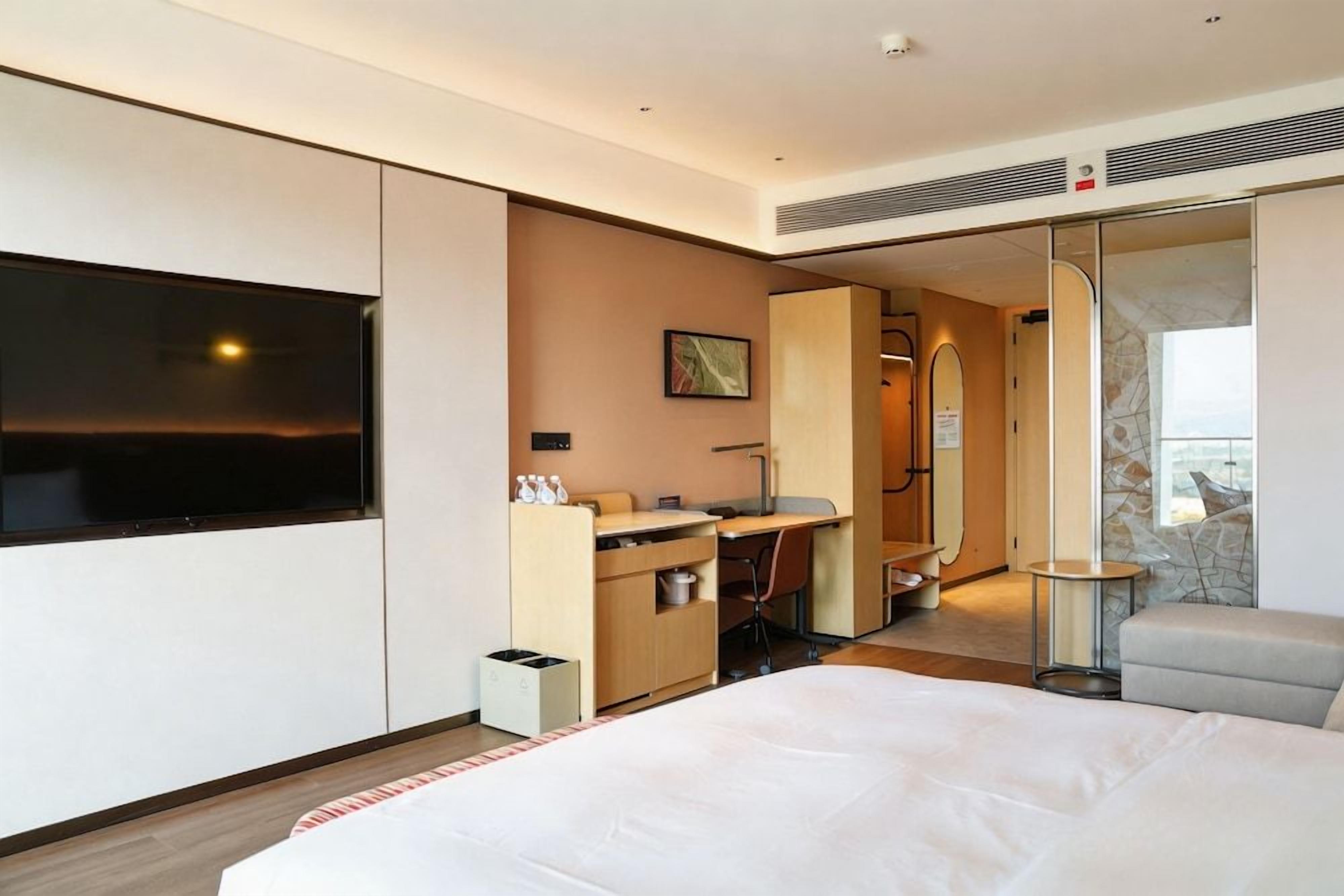 Quarto de hotel moderno com uma cama grande, televisão de tela plana, escrivaninha com cadeira, uma mesinha e banheiro com chuveiro e vaso sanitário.