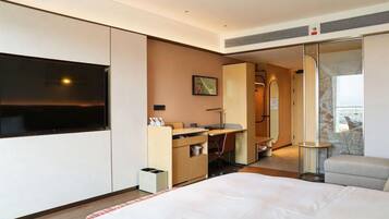 Quarto de hotel moderno com uma cama grande, televisão de tela plana, escrivaninha com cadeira, uma mesinha e banheiro com chuveiro e vaso sanitário.