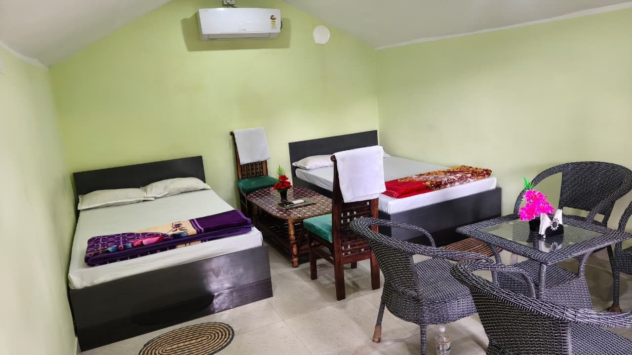 Quarto de hotel com duas camas, uma mesinha, cadeiras de vime e ar condicionado fixado na parede.