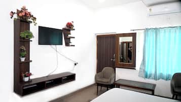 Quarto de hotel com TV de tela plana na parede, prateleira de madeira com plantas em vasos, cama, cadeira e janela com cortinas.
