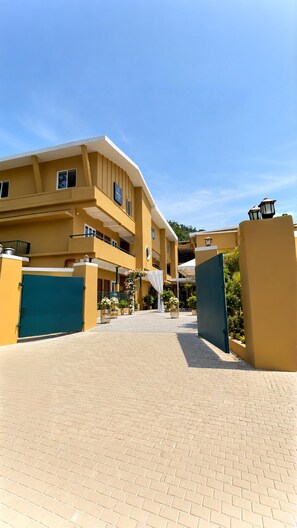 Entrada de hotel com um caminho pavimentado até um portão, um edifício de vários andares com sacadas, e céu claro.