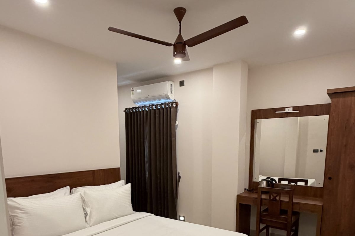 Quarto de hotel com cama, escrivaninha, cadeira, ventilador de teto e ar condicionado.