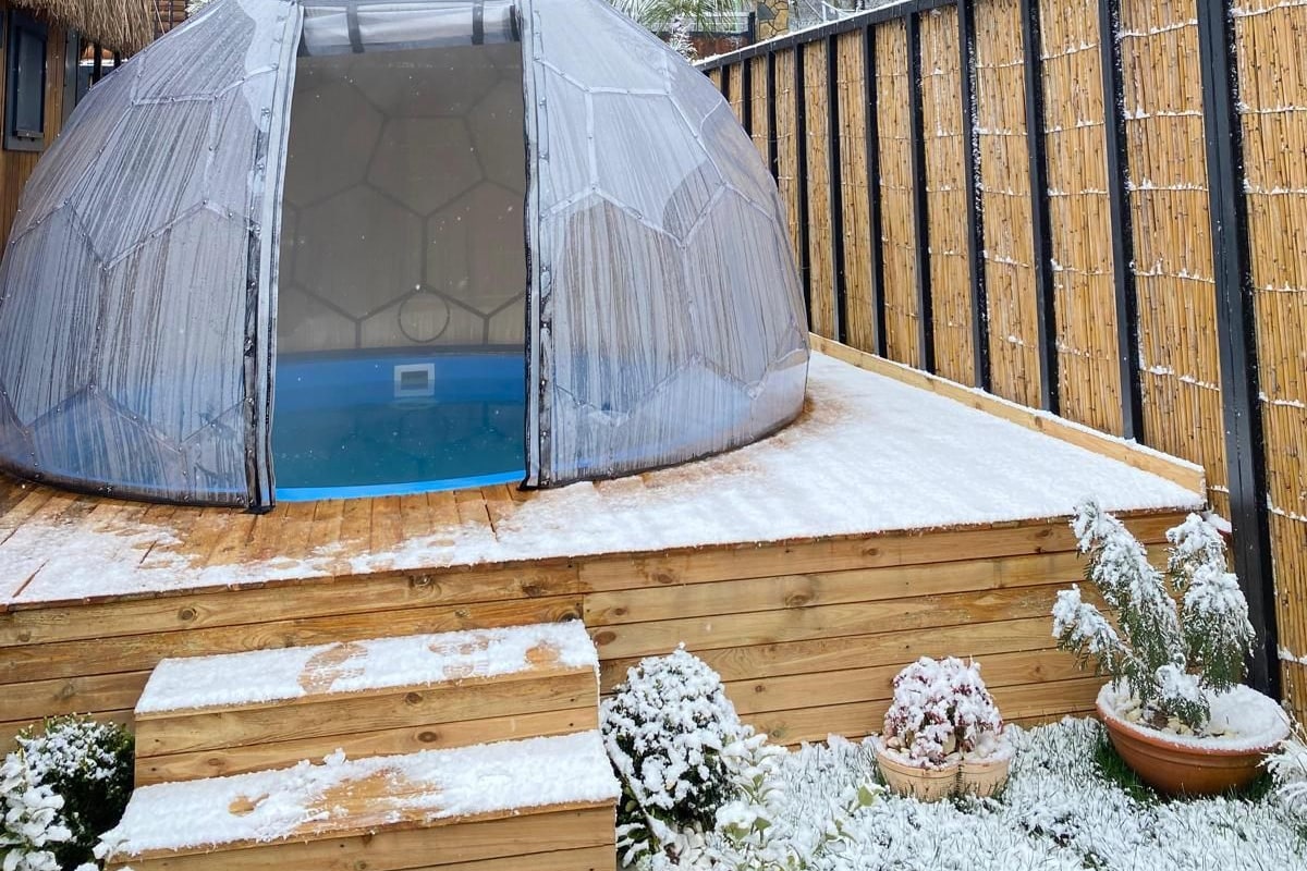 Estrutura em forma de cúpula geodésica com entrada visível, rodeada por um deck de madeira e terreno coberto de neve.
