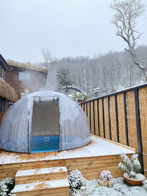 Estrutura em forma de cúpula geodésica com entrada visível, rodeada por um deck de madeira e terreno coberto de neve.