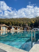 Piscina cristalina com espreguiçadeiras e cabanas com telhado de palha, emoldurada por uma encosta arborizada.