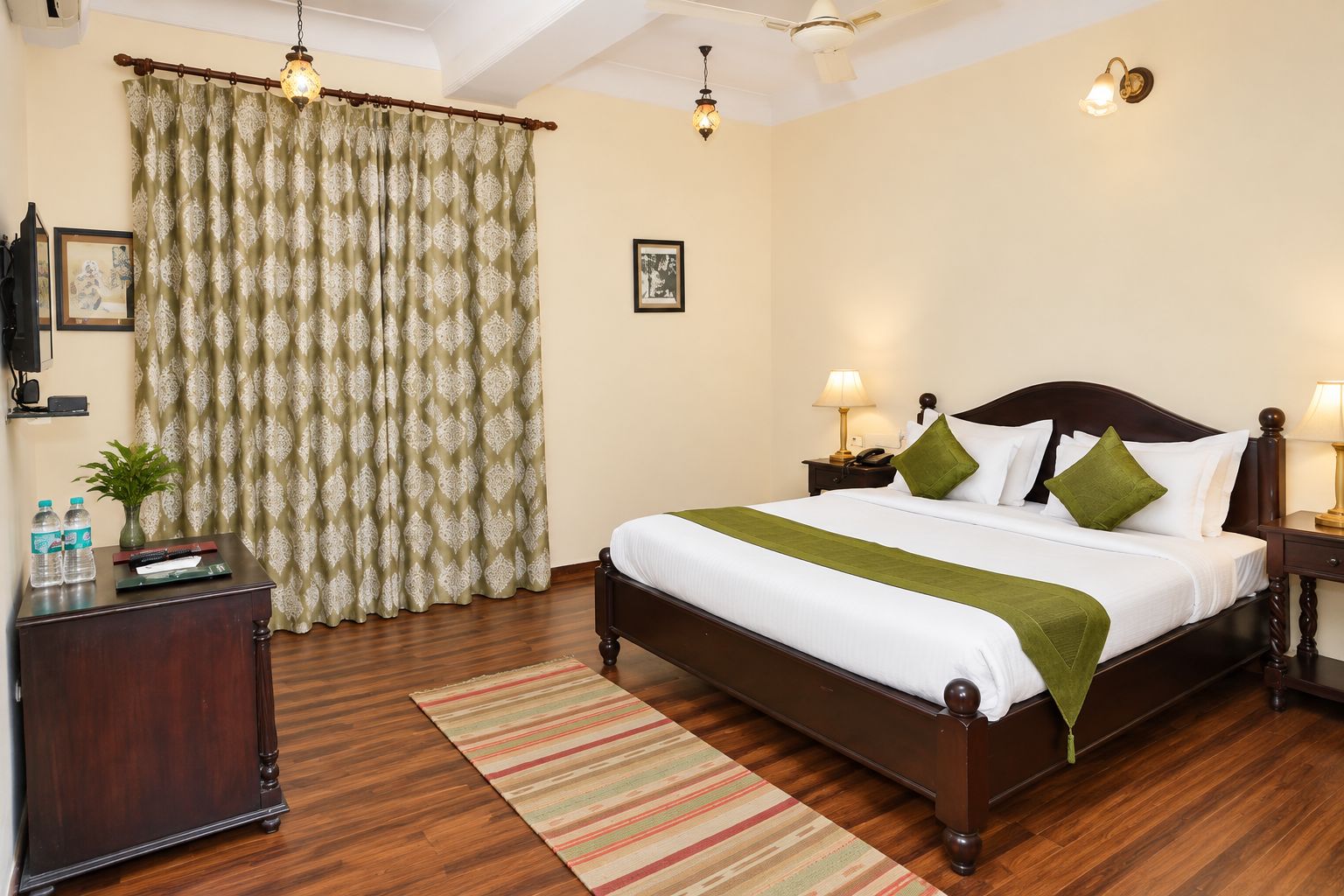 Quarto de hotel com cama grande, abajures, mesa de cabeceira de madeira, escrivaninha com um vaso e garrafas, e cortina estampada.