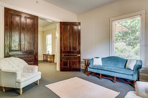 Uma sala de estar com um sofá azul, uma poltrona branca, uma porta de madeira e uma janela com persianas.