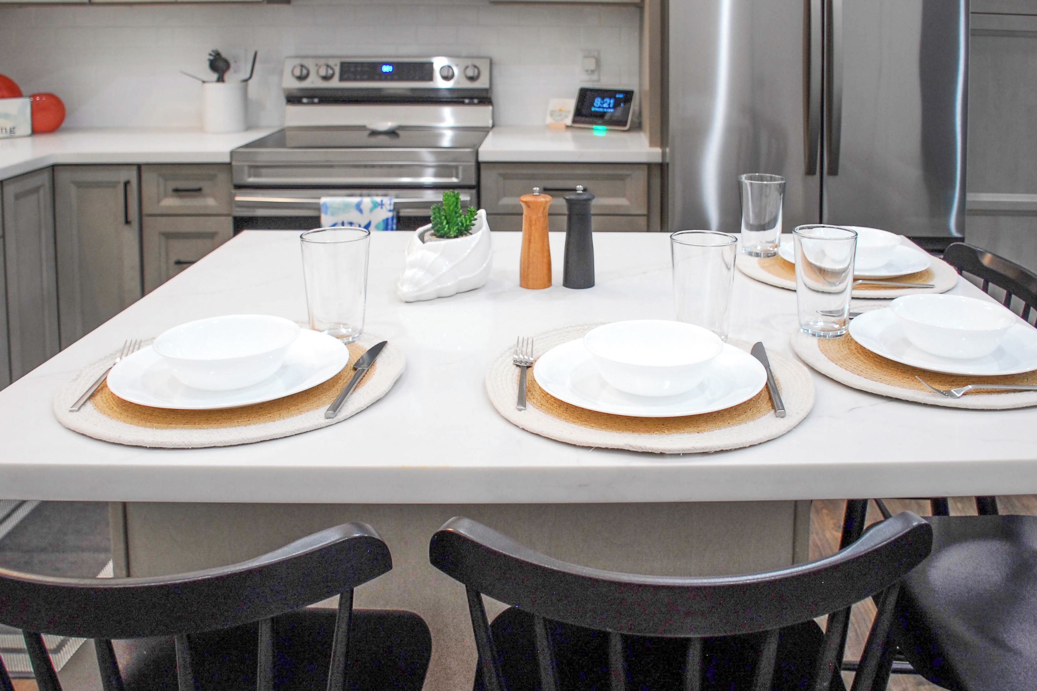 Uma mesa de jantar posta com pratos brancos, talheres e copos de vidro transparente, com uma pequena planta em vaso e dois saleiros e pimenteiros sobre a mesa.