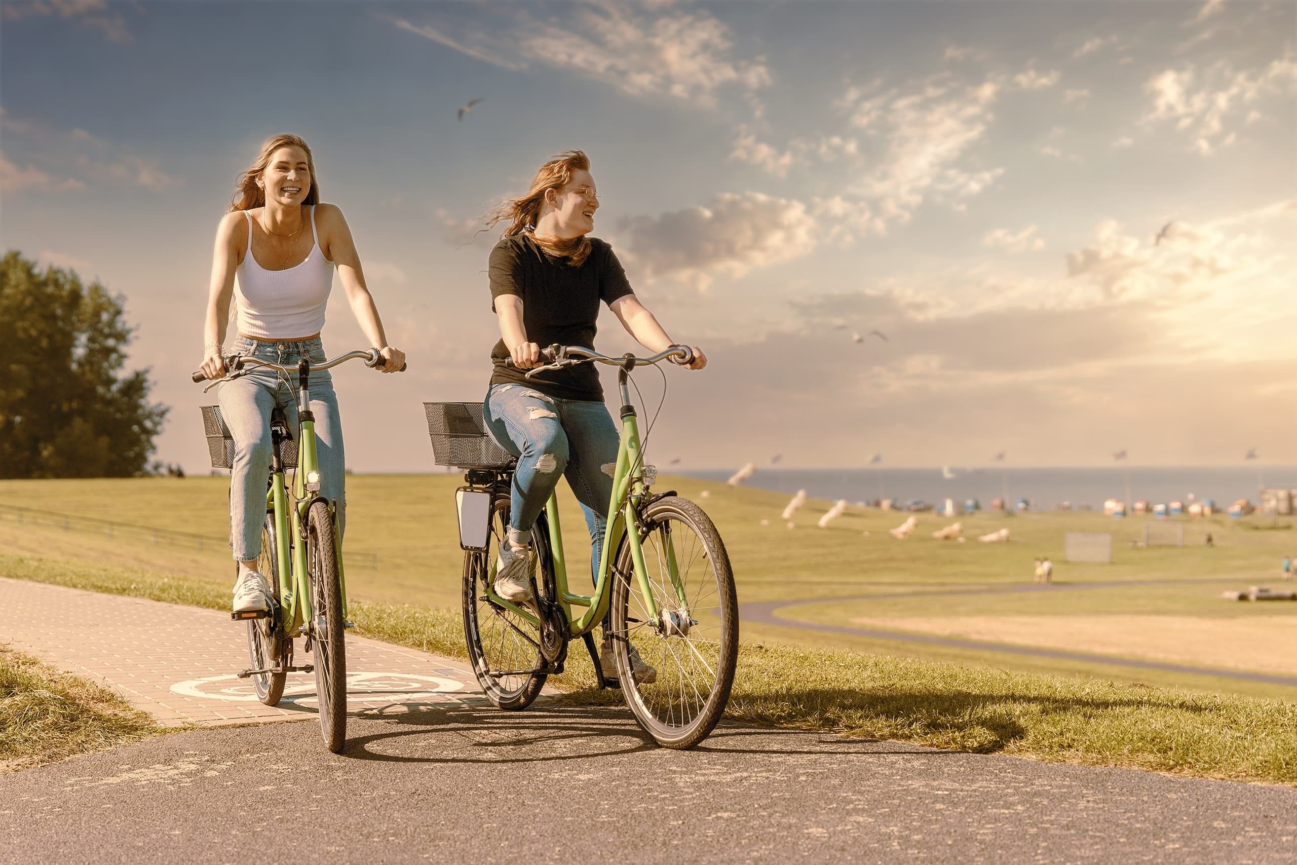Duas pessoas andando de bicicleta em um caminho pavimentado, com um campo gramado e céu azul ao fundo.