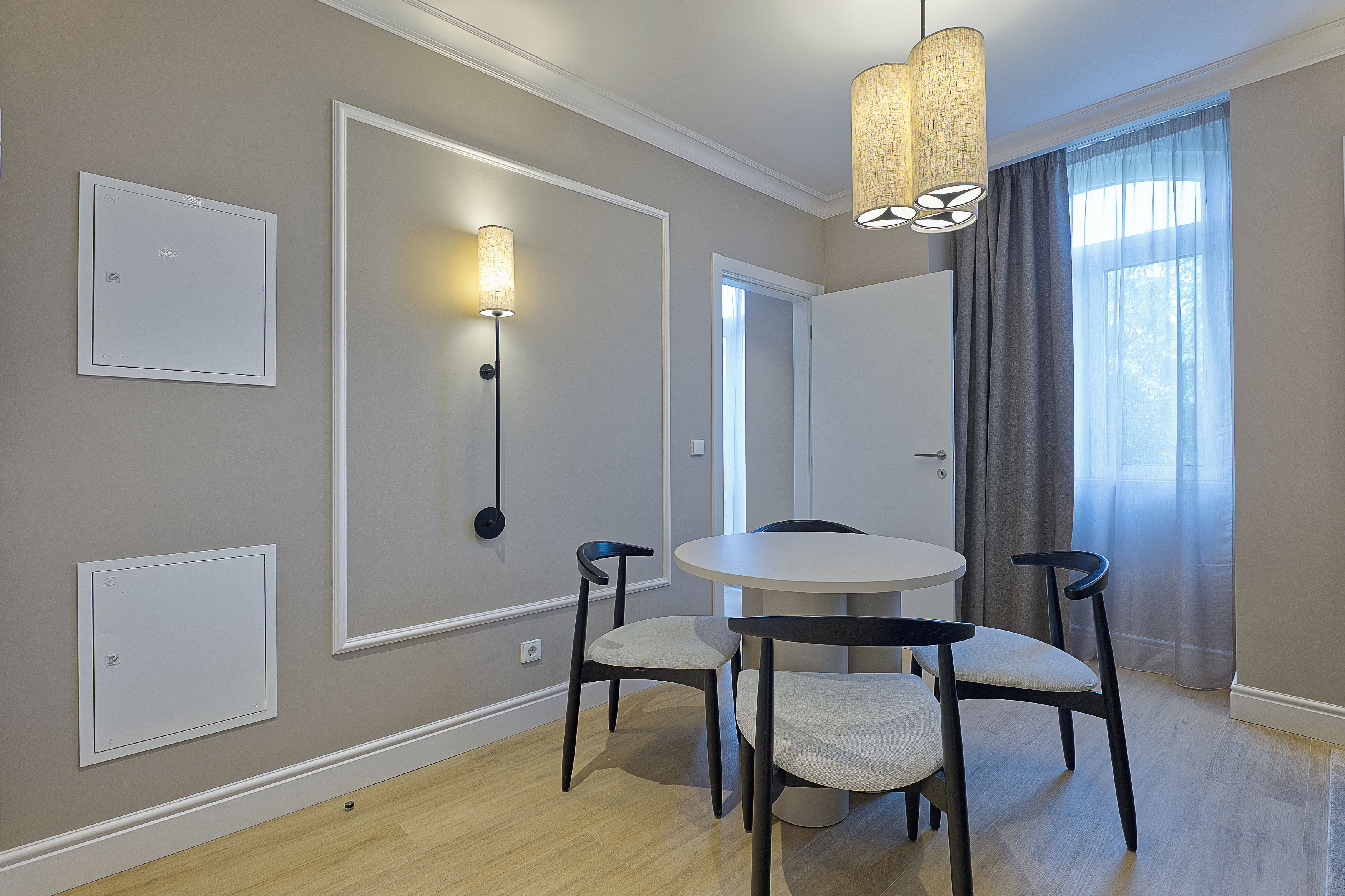 Uma área de refeições com uma mesa redonda e quatro cadeiras, uma luminária fixada na parede e uma porta que dá para outro cômodo.