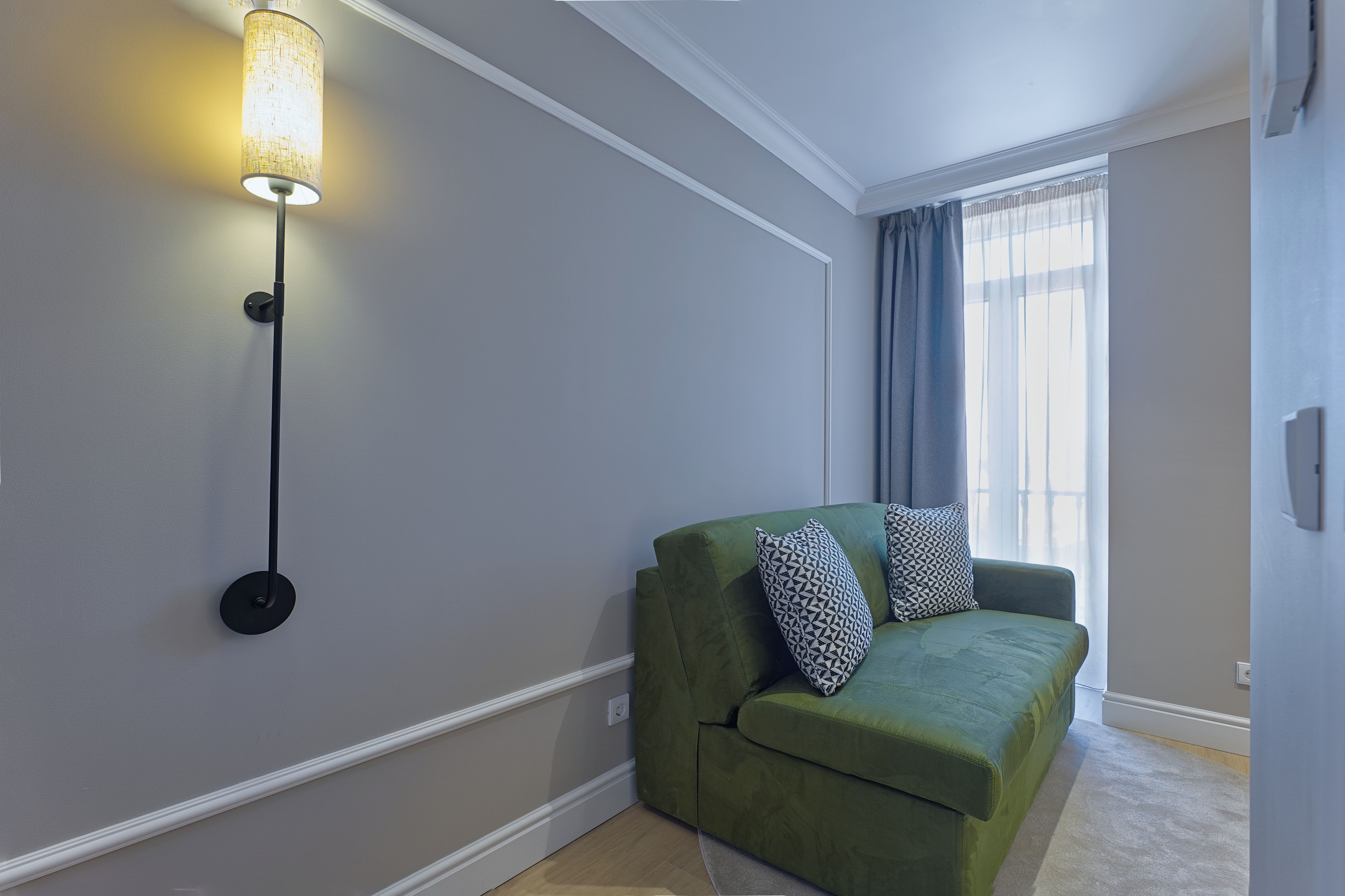 Quarto com um sofá verde, duas almofadas estampadas, um abajur alto e uma janela com cortinas.