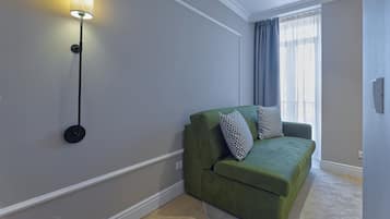 Quarto com um sofá verde, duas almofadas estampadas, um abajur alto e uma janela com cortinas.