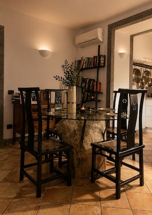 Uma área de refeições com uma mesa de vidro rodeada por cadeiras de madeira, um vaso de planta e uma estante com livros e objetos decorativos.