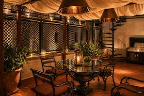 Área de refeições ao ar livre com mesa de vidro, cadeiras de metal, plantas em vasos e estação de churrasco.