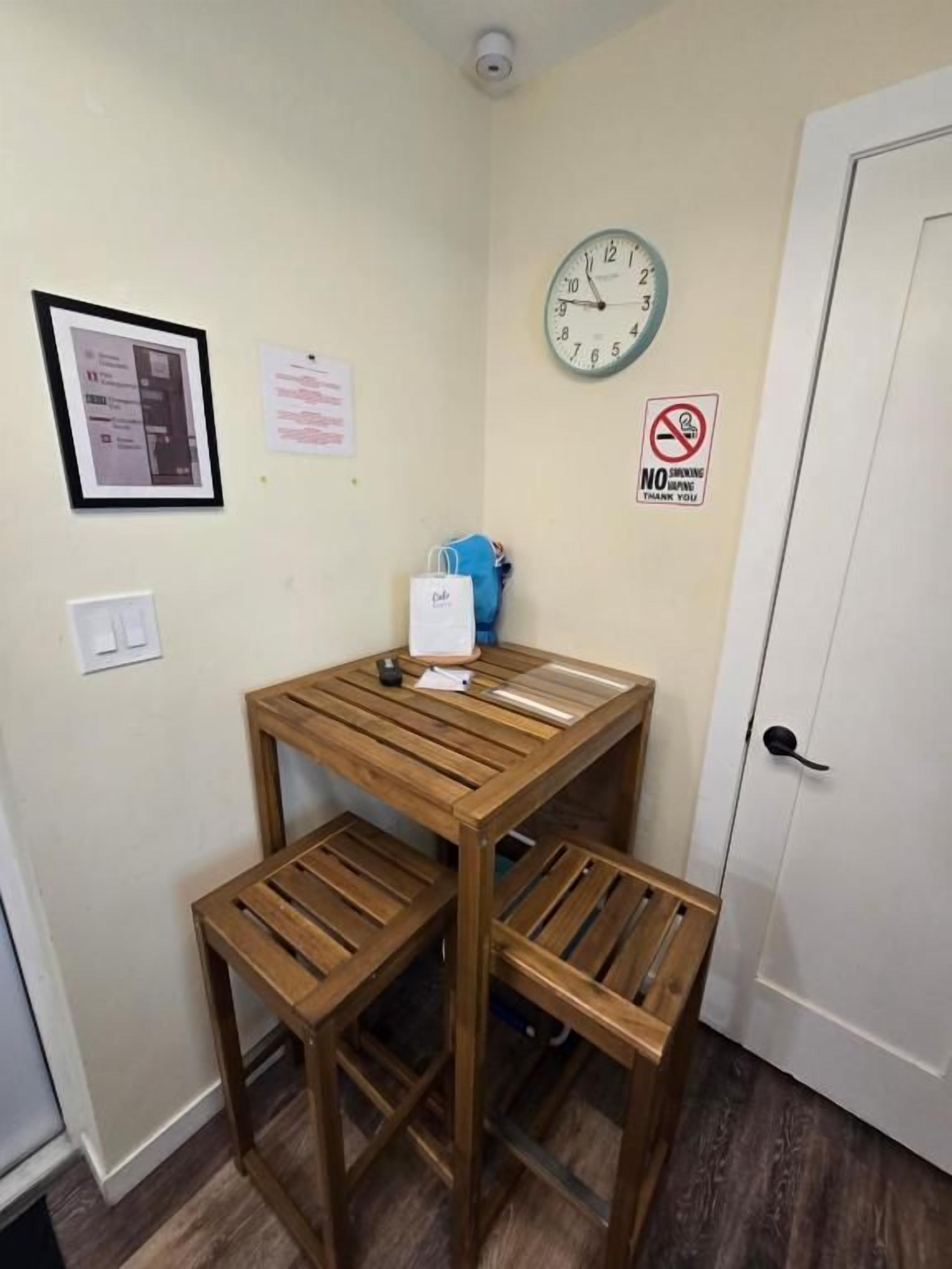 Um quarto pequeno com uma mesa de madeira e duas cadeiras, um relógio na parede e uma porta.