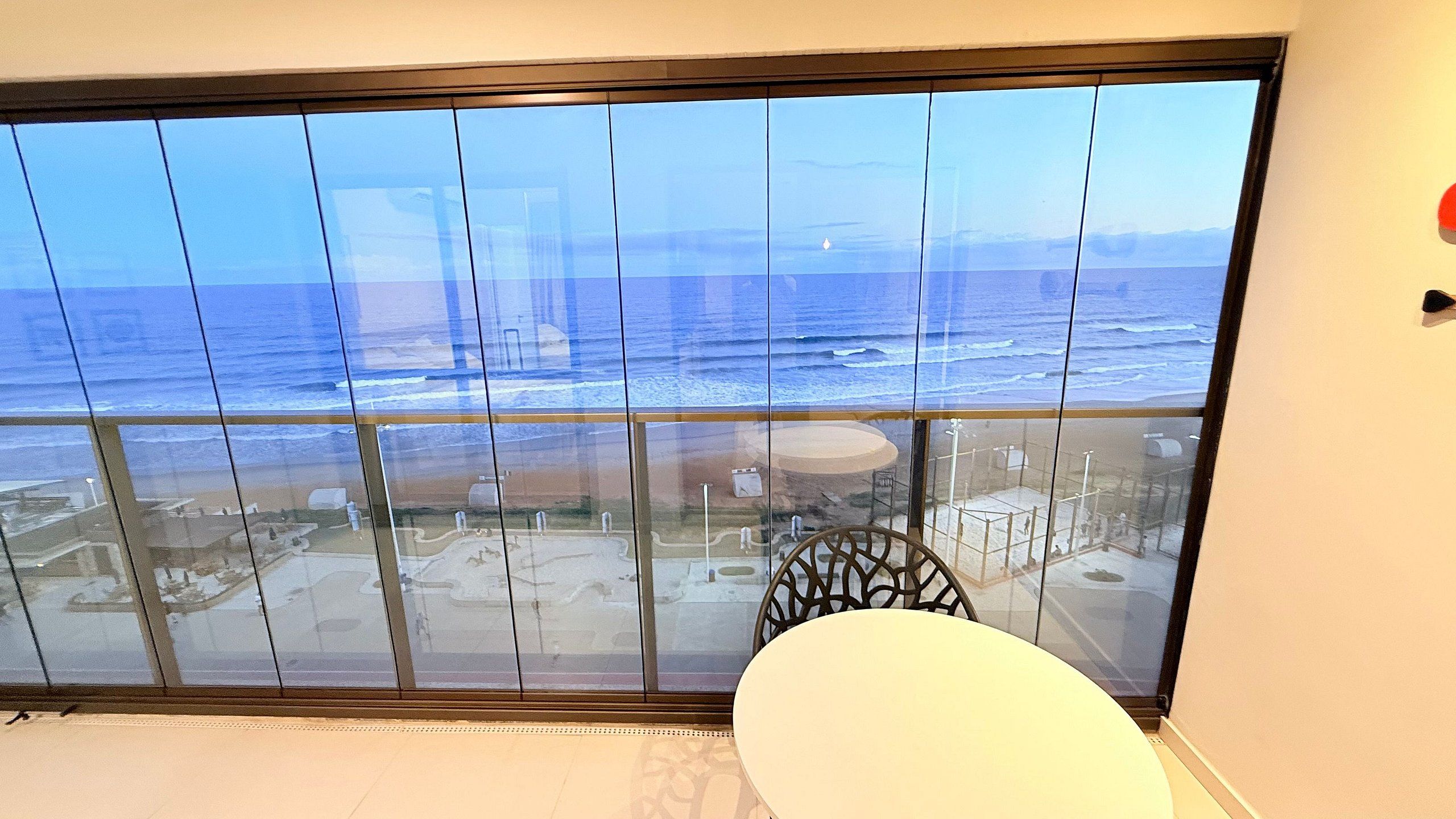 Quarto com grande janela com vista para o oceano, uma cadeira branca e uma pequena mesa.