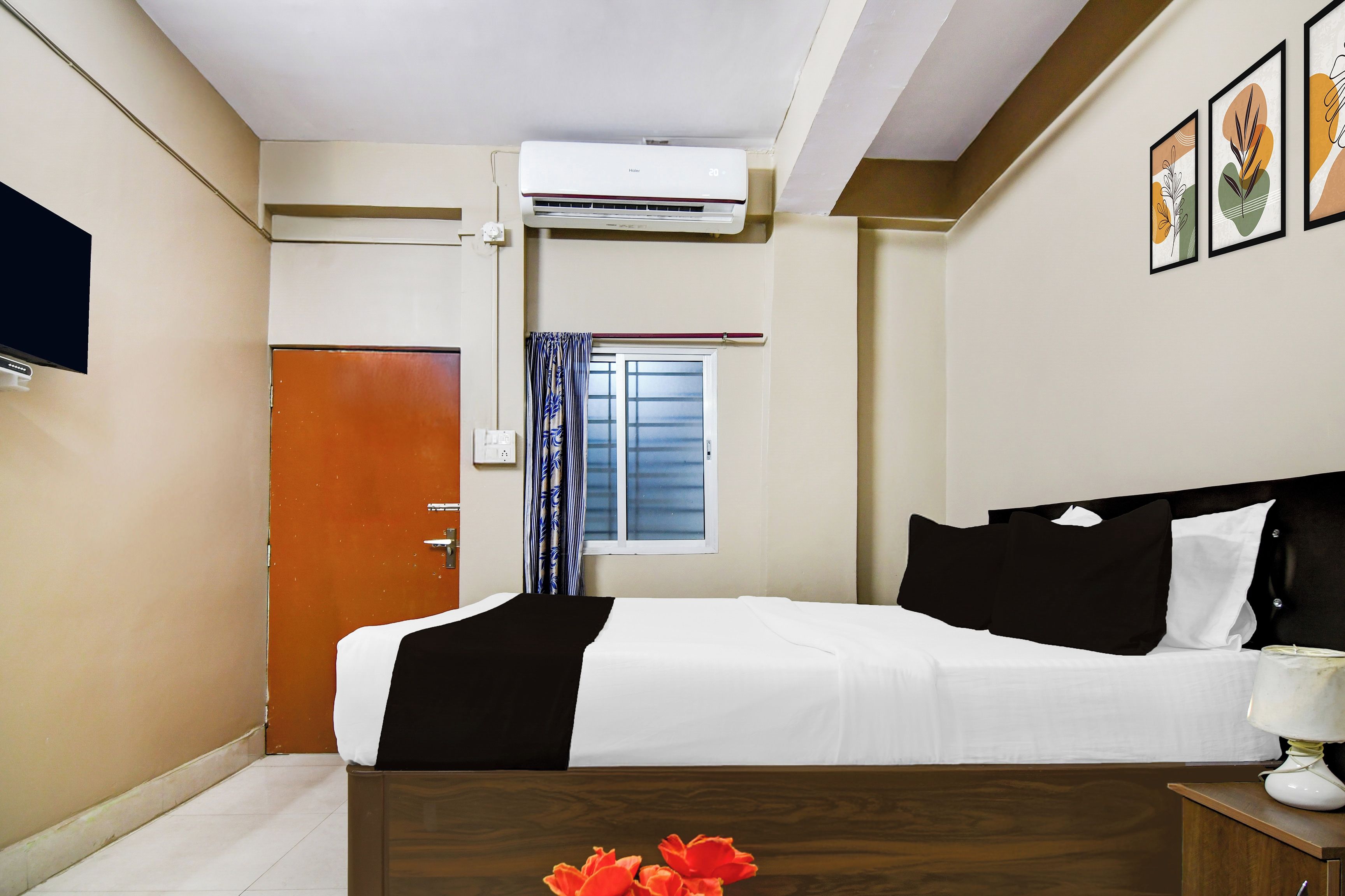 Quarto de hotel com cama, mesa de cabeceira, luminária, aparelho de ar condicionado, janela com cortinas e porta.