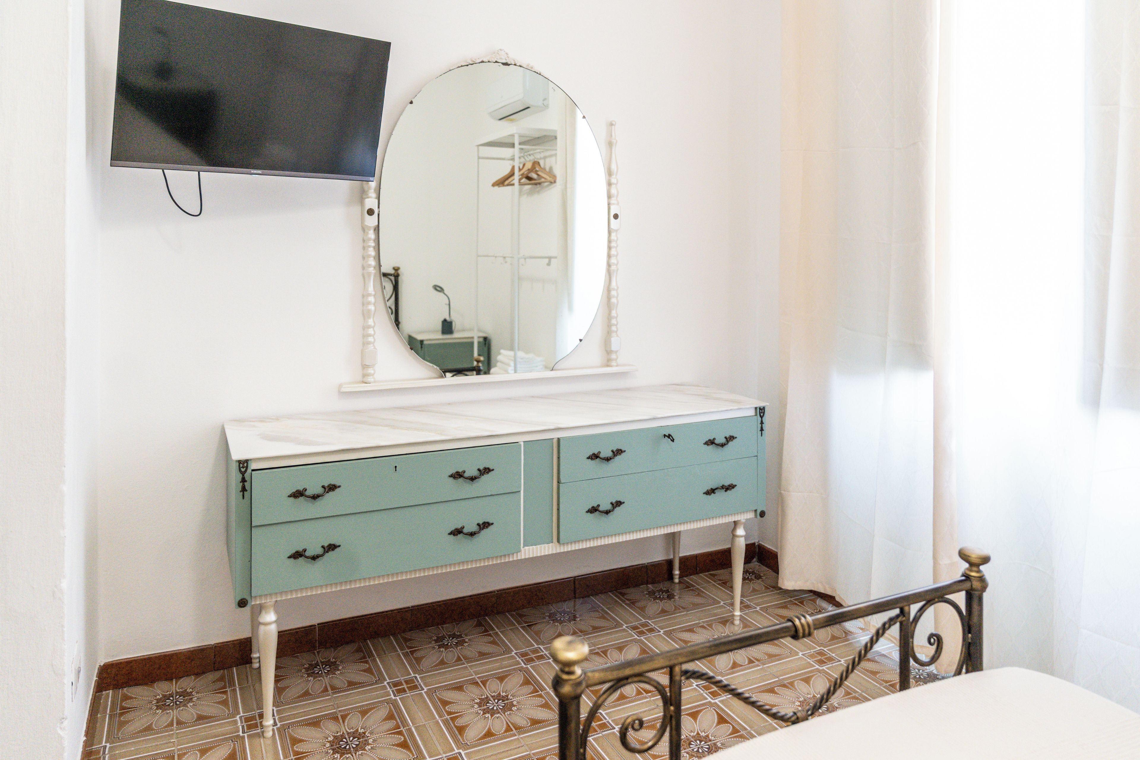 Quarto com cama, cómoda com gavetas, espelho e televisão na parede.