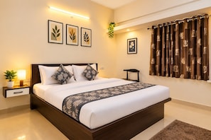 Quarto de hotel com uma cama grande, mesa de cabeceira com abajur, uma cadeira pequena, planta em vaso e uma janela com cortinas.