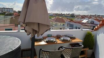 Varanda com mesa posta para refeições, cadeiras, planta em vaso e vista da paisagem urbana.