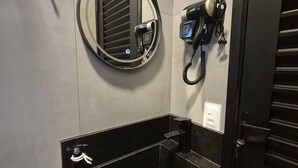Casa de banho com bancada preta, espelho redondo, secador de cabelo e suporte para toalhas.