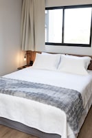 Quarto com cama grande, mesa de cabeceira, luminária e janela com cortinas.