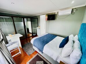 Quarto de hotel moderno com cama, escrivaninha, televisão e porta deslizante para o banheiro.