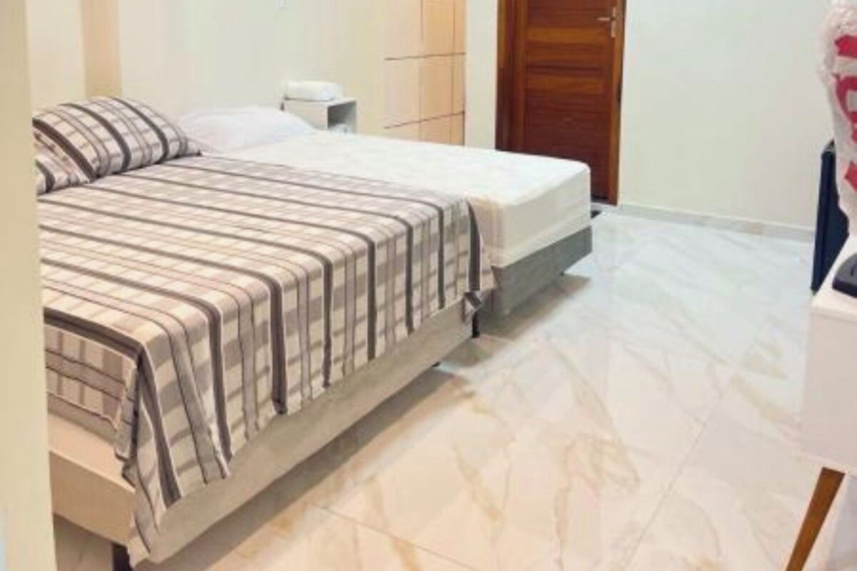 Quarto com cama, mesa de cabeceira, porta de madeira e parede branca.