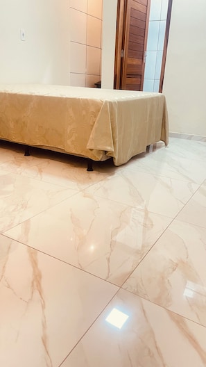 Quarto com cama coberta por um edredom bege, chão de azulejos com padrão marmorizado e porta de madeira ao fundo.