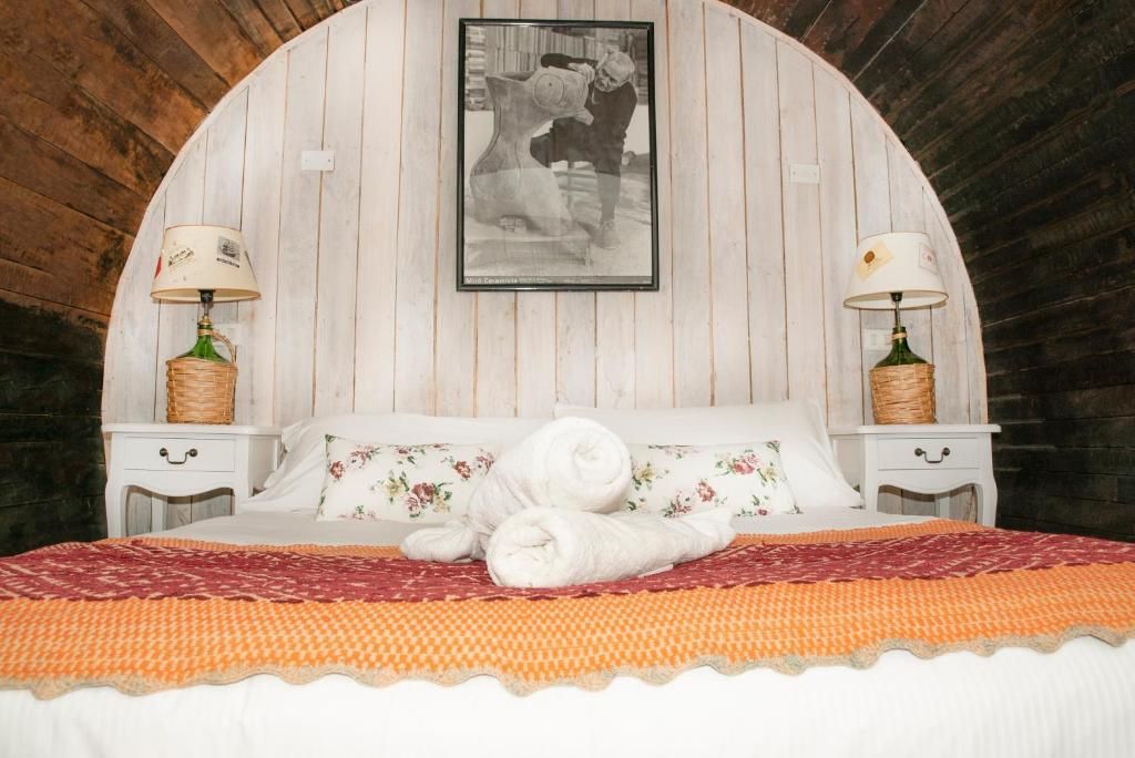 Uma cama com edredom branco, duas almofadas estampadas com motivos florais e uma toalha enrolada. Há duas mesas de cabeceira com luminárias e cestos. Uma moldura com uma foto está pendurada acima da cama.