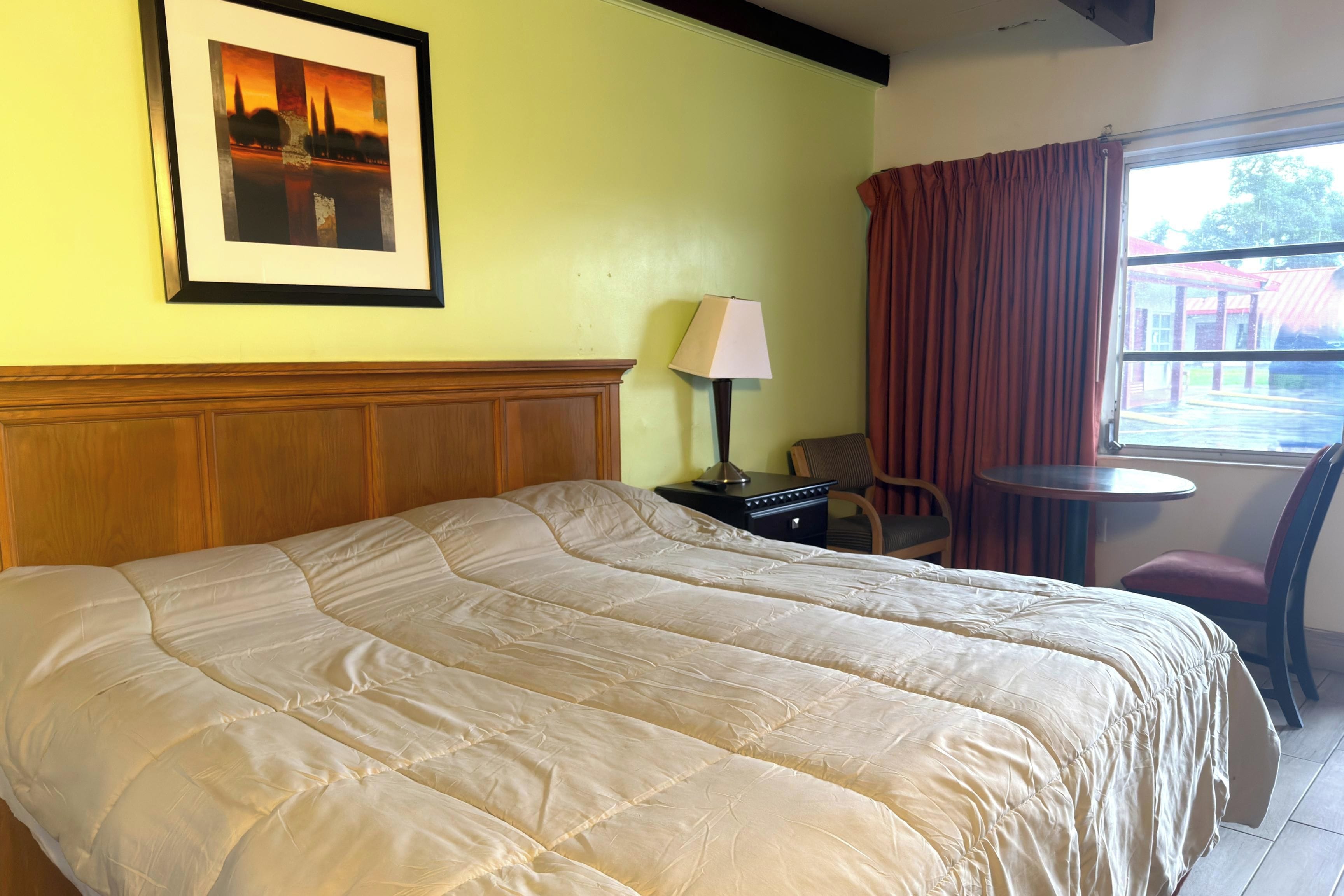 Quarto de hotel com cama grande, mesa de cabeceira, luminária, cadeira, mesinha e janela com cortinas.