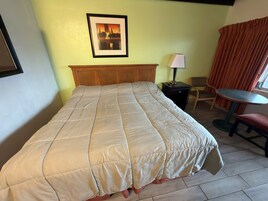 Quarto de hotel com uma cama grande, um quadro na parede, uma mesa pequena, uma cadeira e uma janela com cortinas.