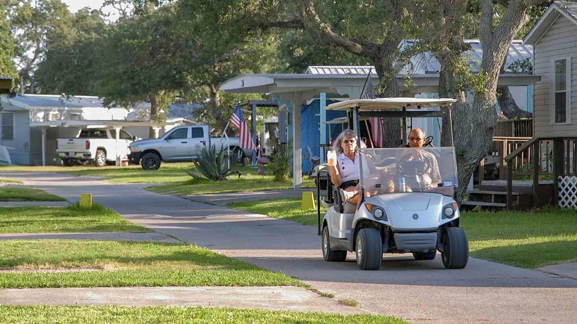 Rua residencial com casas, uma caminhonete estacionada e um carrinho de golfe com dois passageiros.