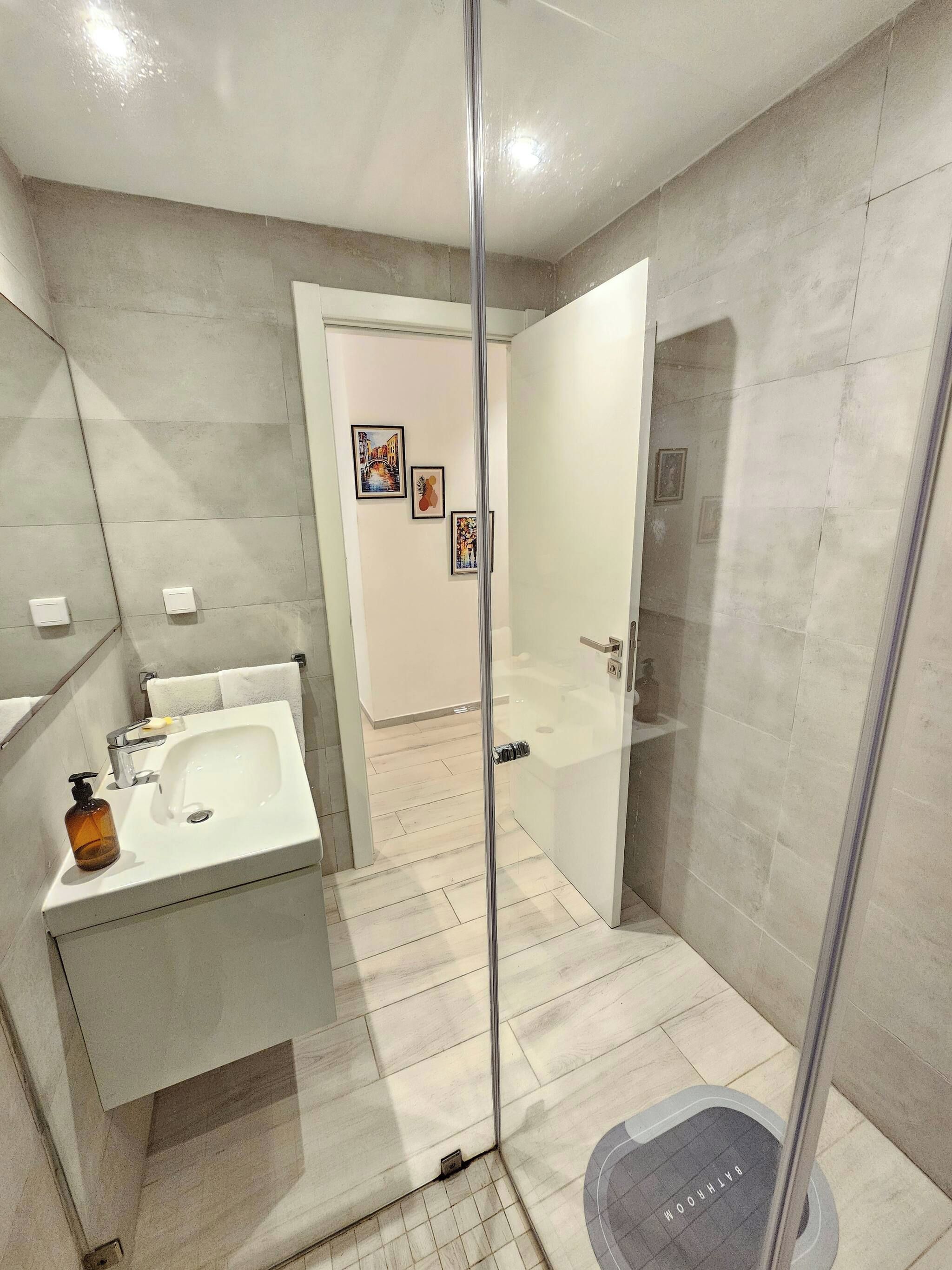 Casa de banho moderna com cabine de chuveiro em vidro, lavatório branco com dispensador de sabão e porta que dá para outro espaço.