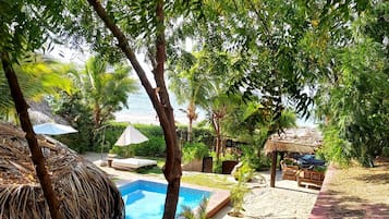 Uma área de piscina com guarda-sol de palha, espreguiçadeiras e vista para a praia.